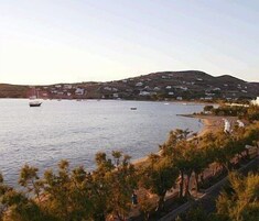 Vista costeira com barcos na água, praia com árvores e um morro com edifícios.