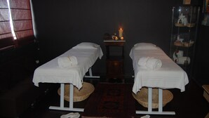 Quarto com duas camas de solteiro, uma mesinha com uma vela e uma prateleira com artigos de higiene pessoal.