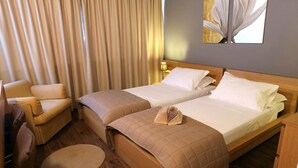 Quarto de hotel com cama, duas cadeiras, uma mesa redonda pequena e um quadro na parede.