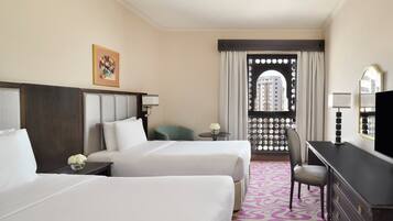 Quarto de hotel com duas camas, uma escrivaninha, uma cadeira, uma mesinha de centro com um vaso, e uma janela com cortinas.