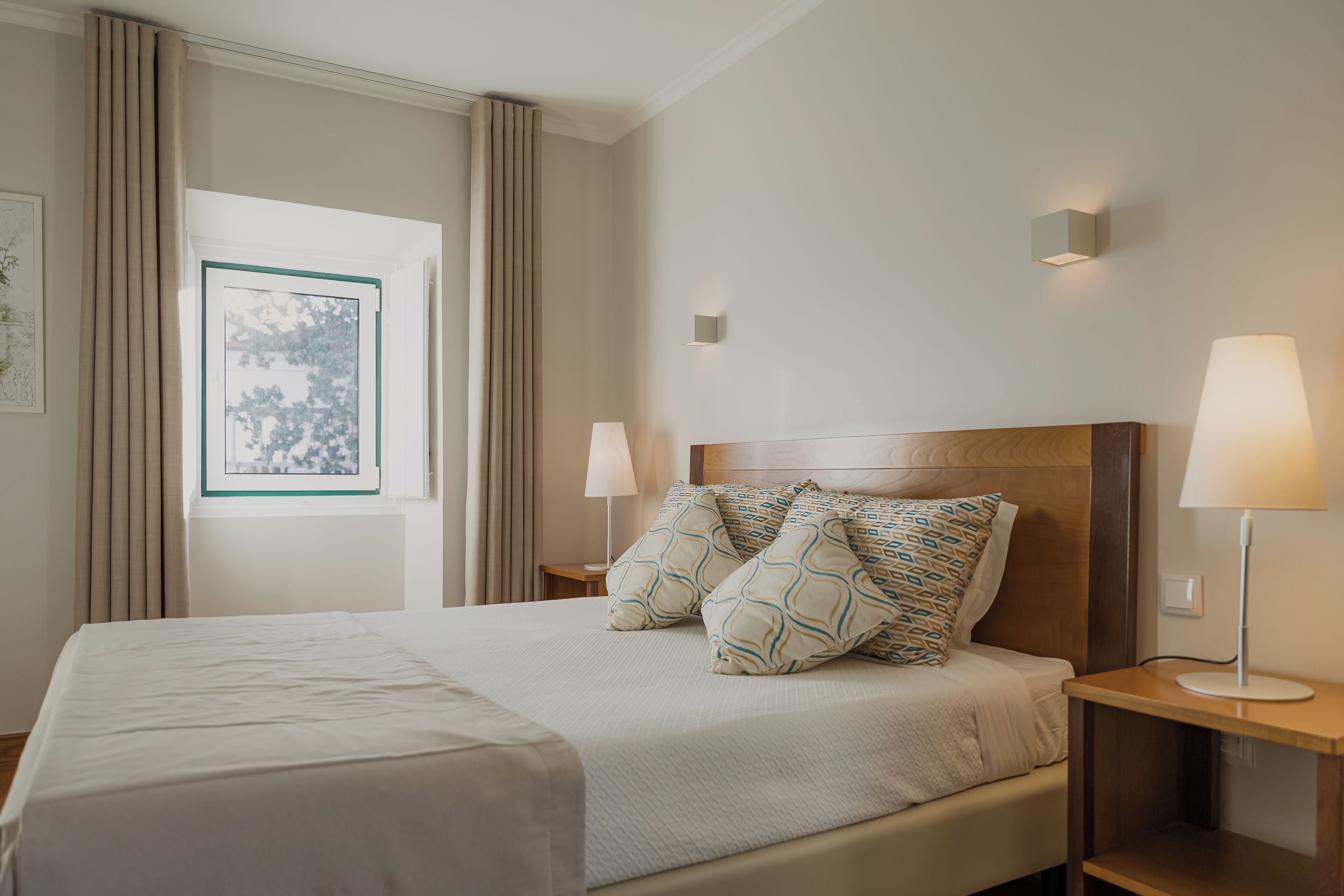 Cama bem arrumada com almofadas estampadas, abajur e janela com cortinas.