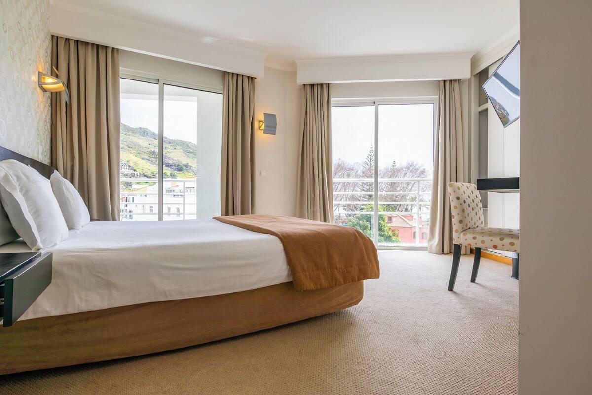 Quarto de hotel com uma cama grande, uma escrivaninha, uma cadeira e vista para o exterior.