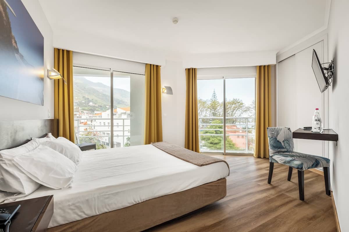 Quarto de hotel moderno com uma cama grande, uma escrivaninha com cadeira e vista da cidade através de amplas janelas.
