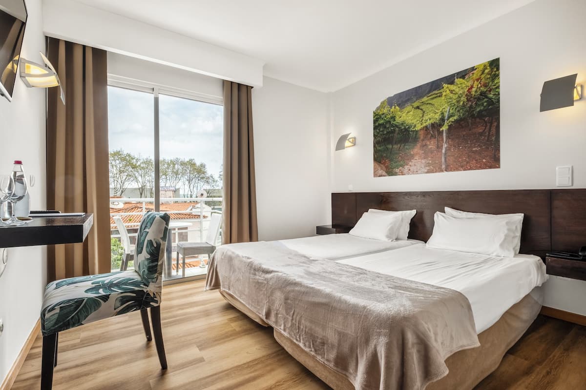 Quarto de hotel com uma cama grande, uma poltrona com padrão de folha, uma mesa com garrafa e copos, e uma pintura de vinhedo na parede.