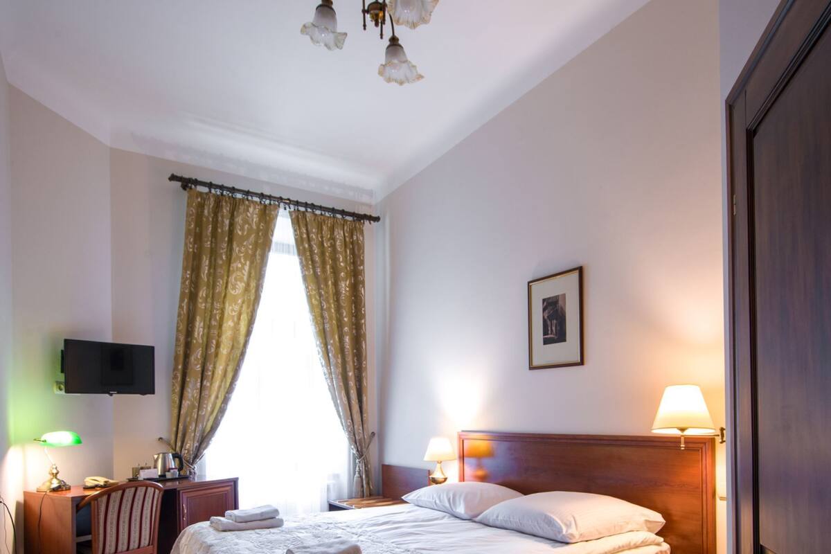 Quarto de hotel com cama, abajures, escrivaninha com cadeira, televisão e janela com cortinas.