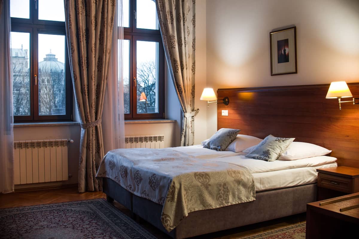 Quarto de hotel com uma cama grande, duas luminárias de cabeceira, uma cabeceira de madeira, um quadro na parede e vista para prédios pela janela.