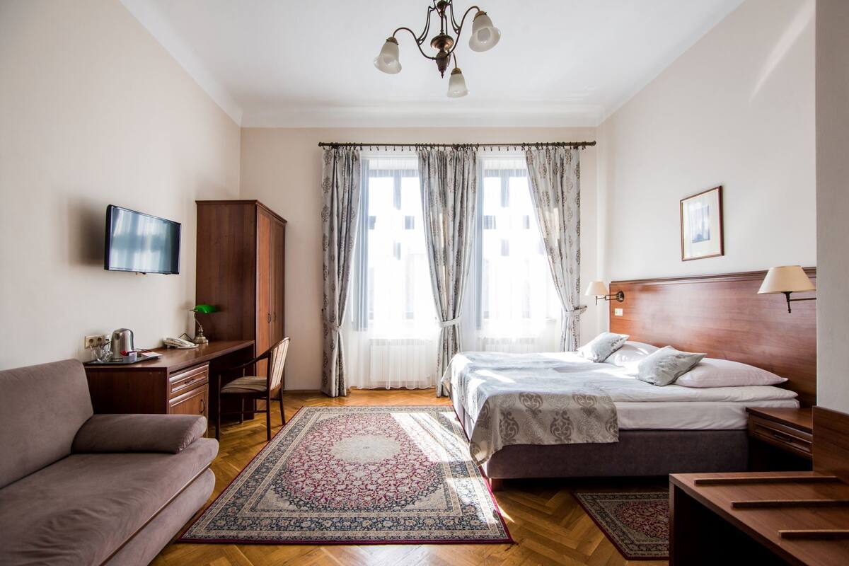 Quarto com cama, escrivaninha, cadeira, televisão e uma grande janela com cortinas.