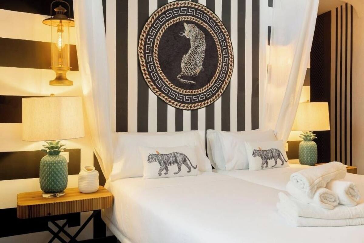 Quarto de hotel com uma cama, duas almofadas com estampas de animais, uma obra de arte circular pendurada na parede, uma mesa de cabeceira com abajur e uma pequena planta em vaso.