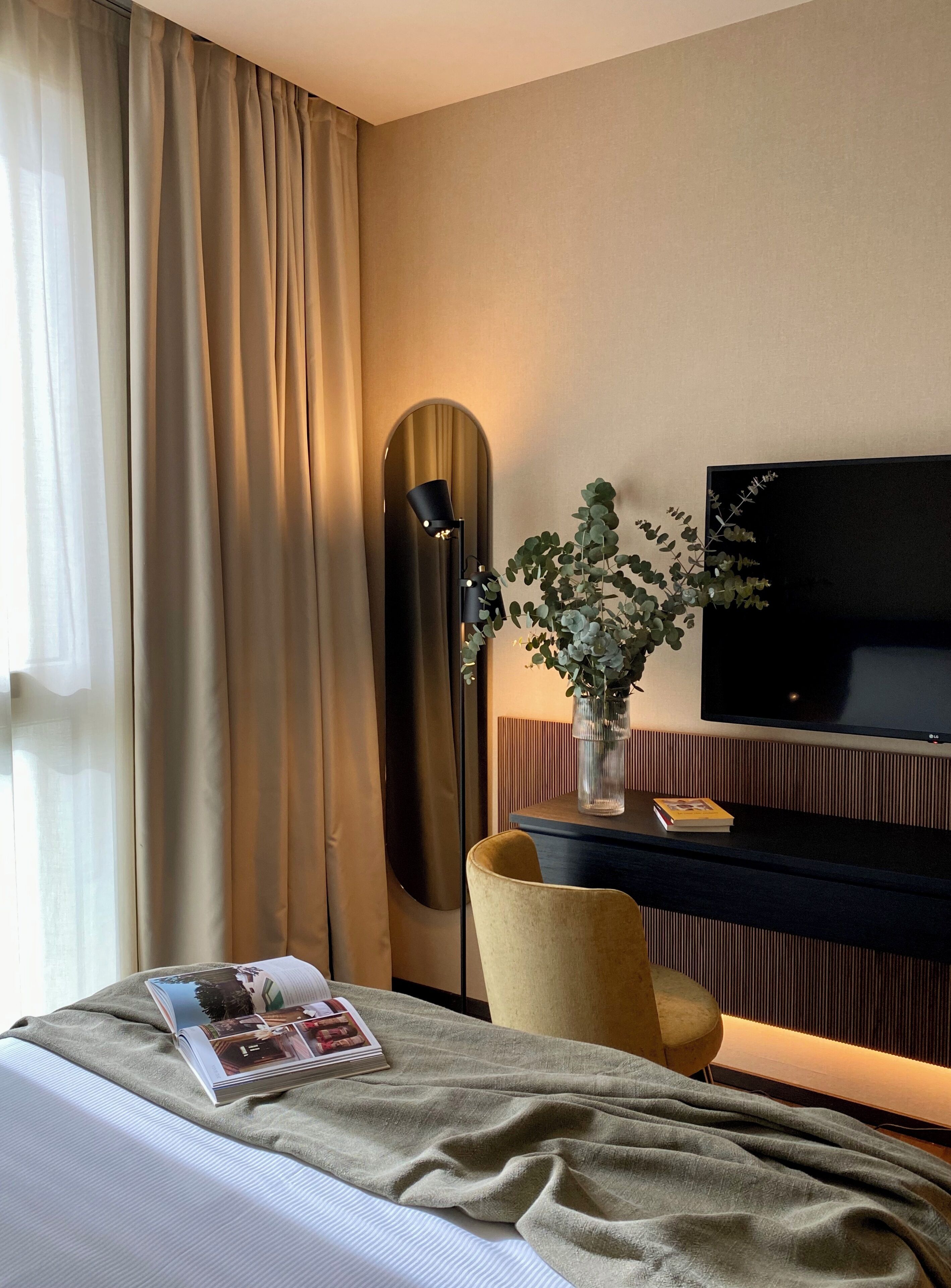 Quarto de hotel com cama, mesa de cabeceira com um livro, televisão e uma planta.