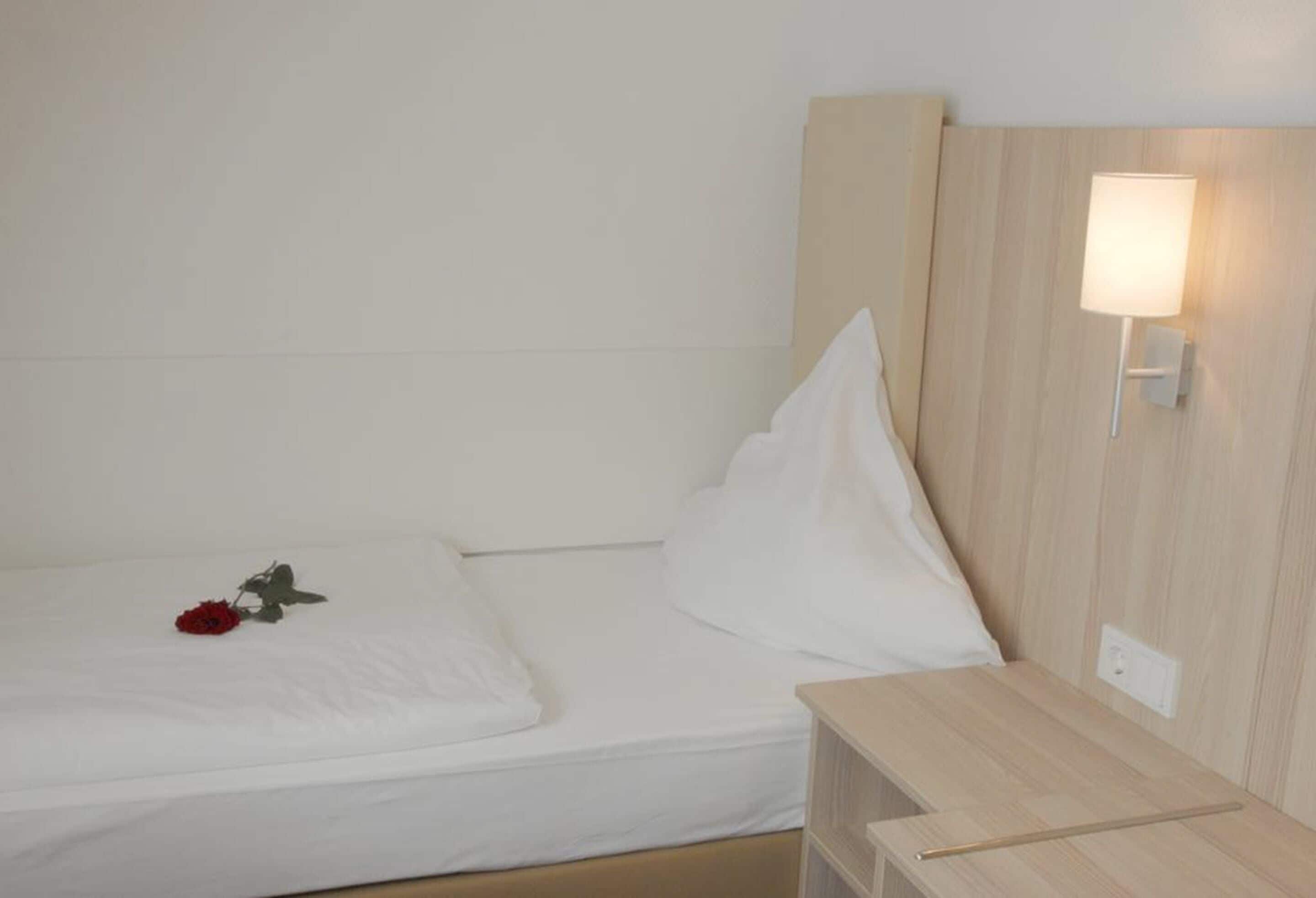 Cama bem arrumada com lençóis brancos, uma única rosa vermelha e uma mesa de cabeceira fixa na parede com abajur.