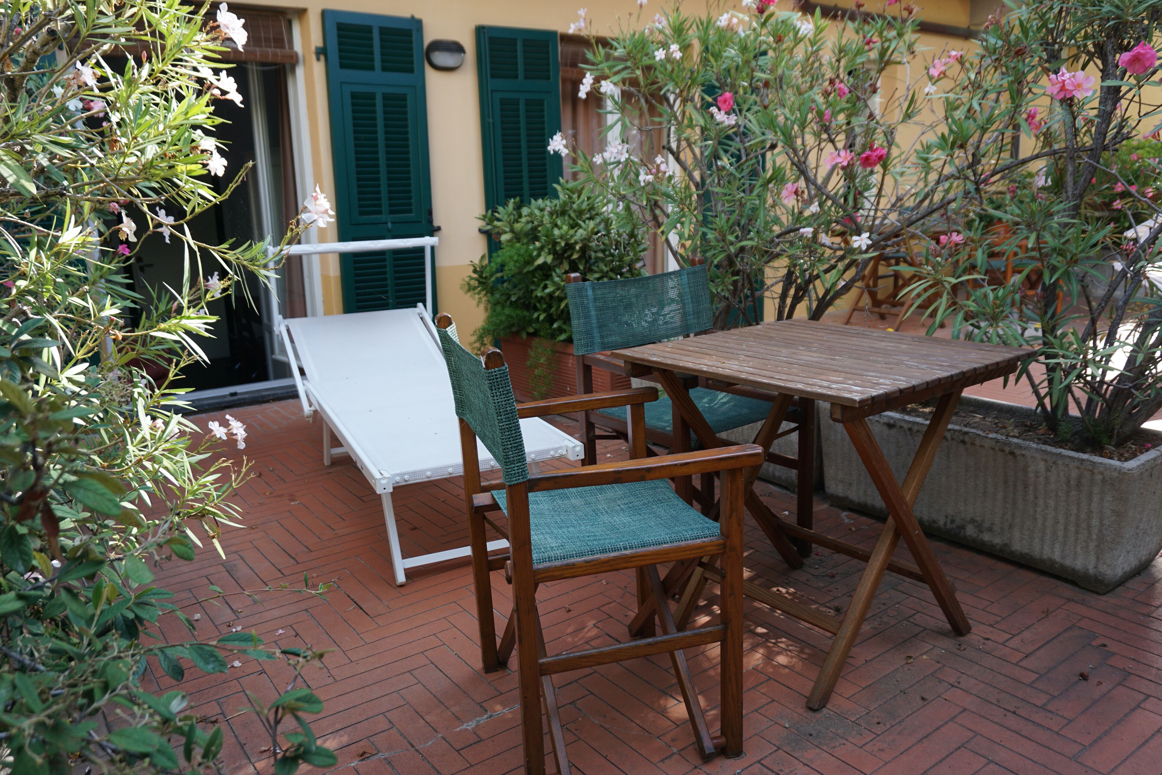 Um pátio externo com mesa e cadeiras de madeira, uma poltrona branca e vasos com plantas.