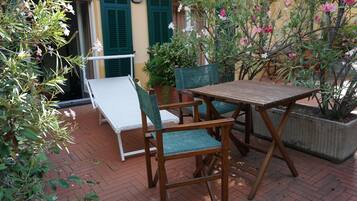 Um pátio externo com mesa e cadeiras de madeira, uma poltrona branca e vasos com plantas.
