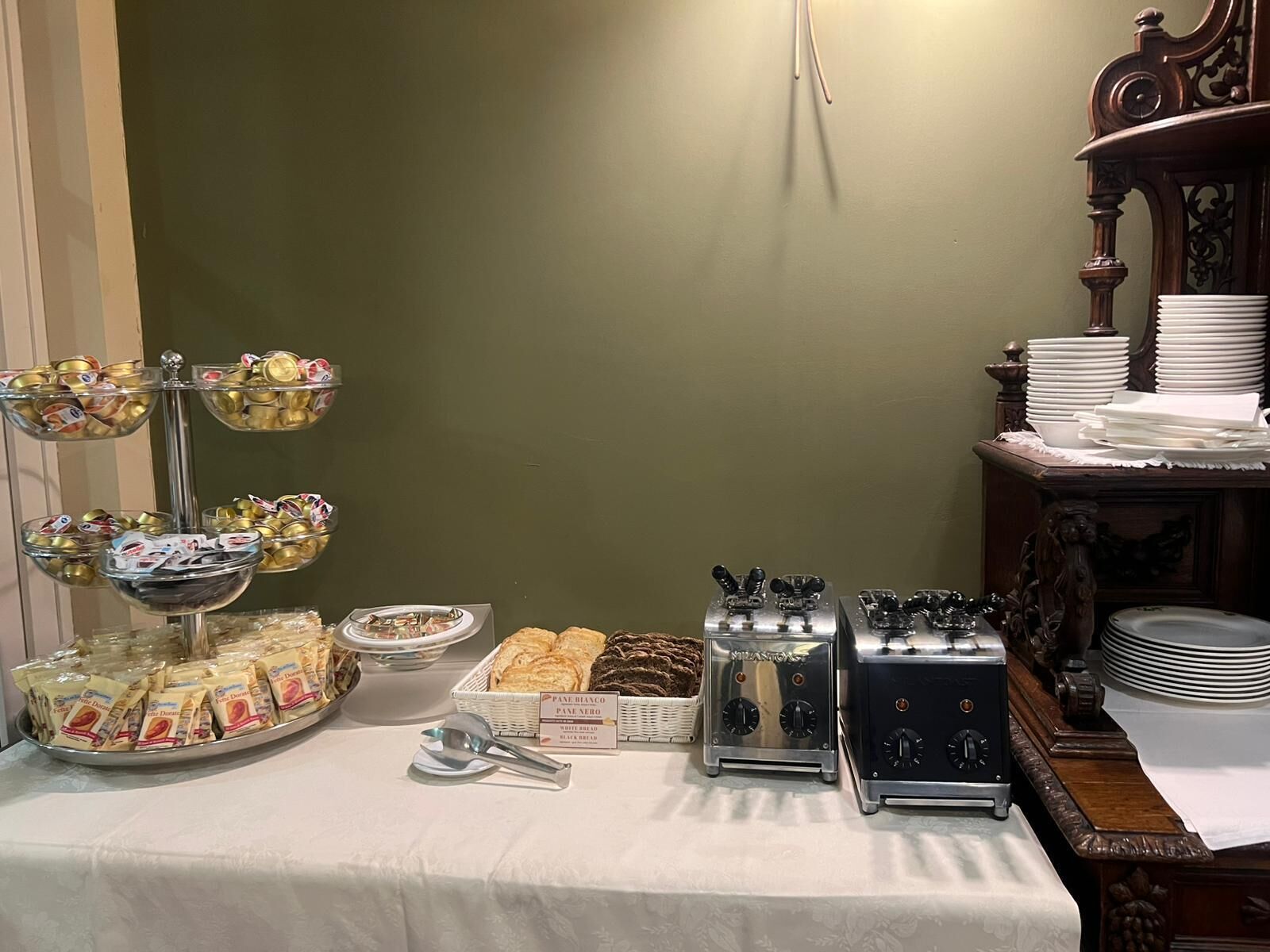 Uma mesa de buffet com diversos alimentos, incluindo doces, pão e acompanhamentos. Há também duas máquinas de pão e pilhas de pratos brancos.