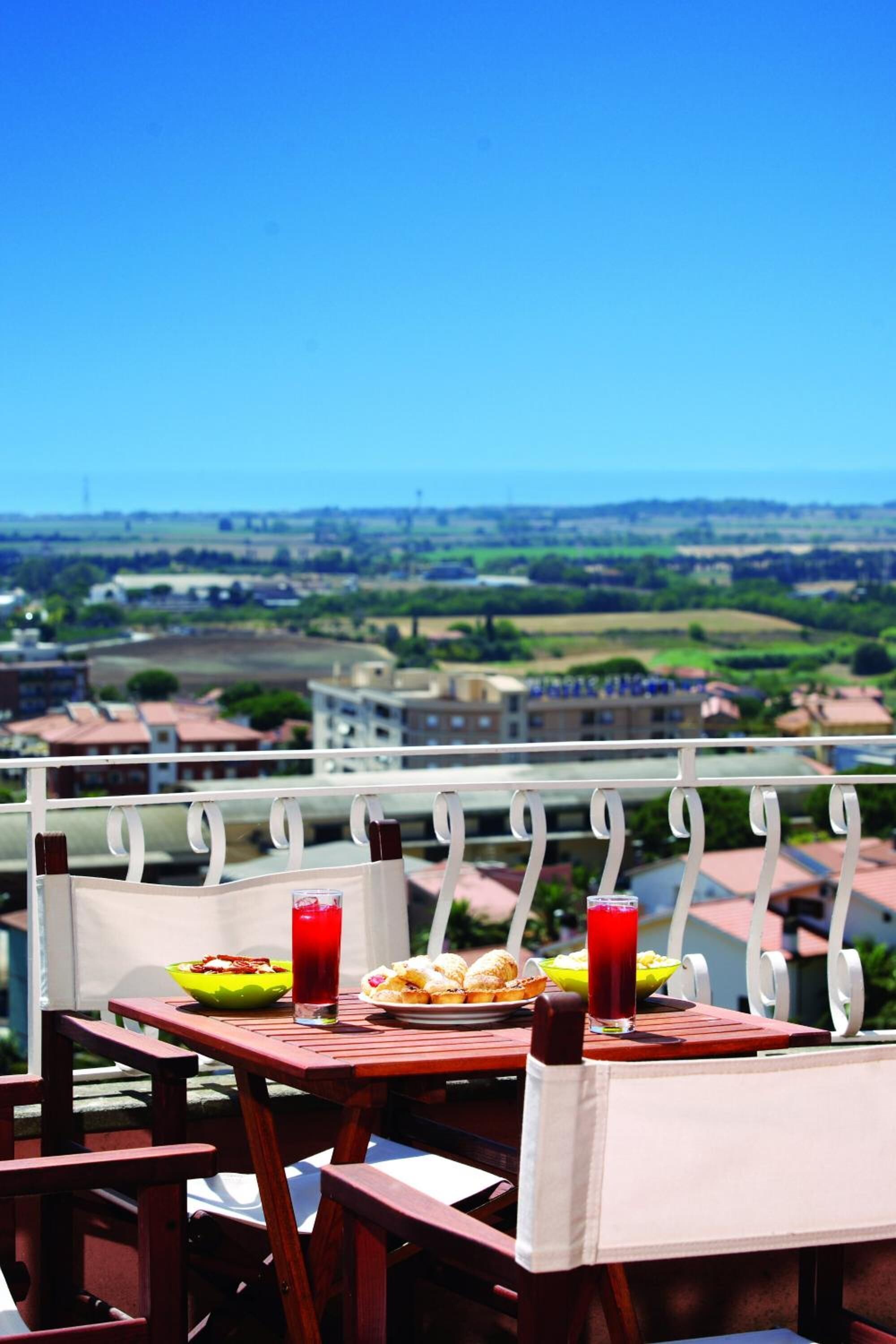 Uma mesa com uma bebida vermelha, um prato de comida e vista para uma cidade com edifícios e áreas verdes.