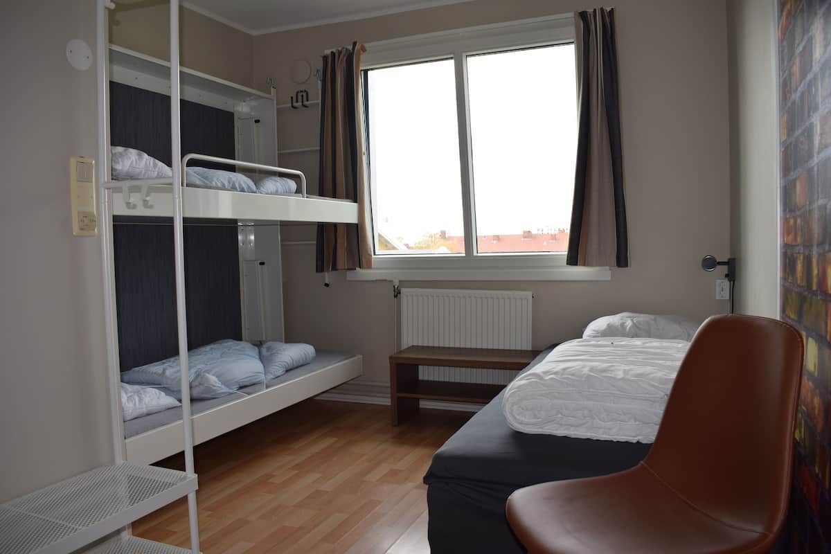 Quarto em dormitório com beliches, uma cama de solteiro, uma cadeira e uma janela com cortinas.
