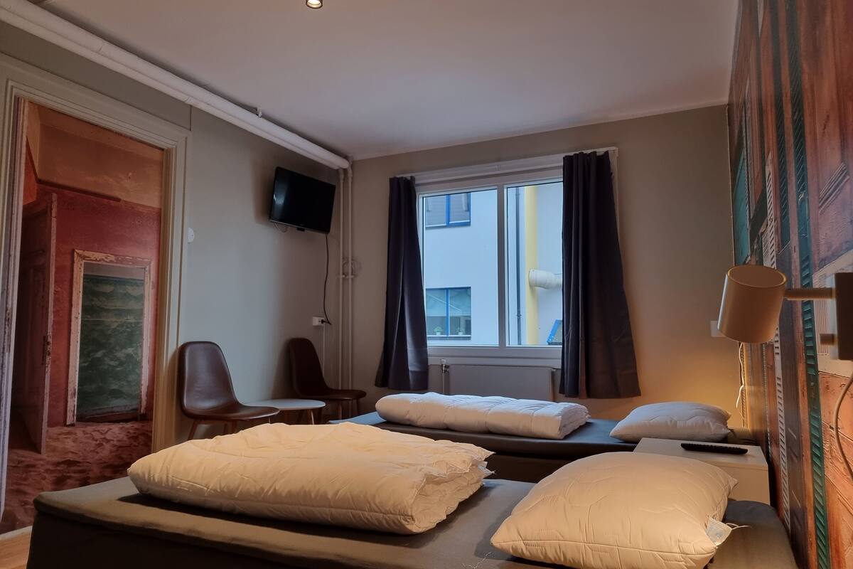 Quarto de hotel com duas camas, televisão fixada na parede, janela com cortinas e uma cadeira.