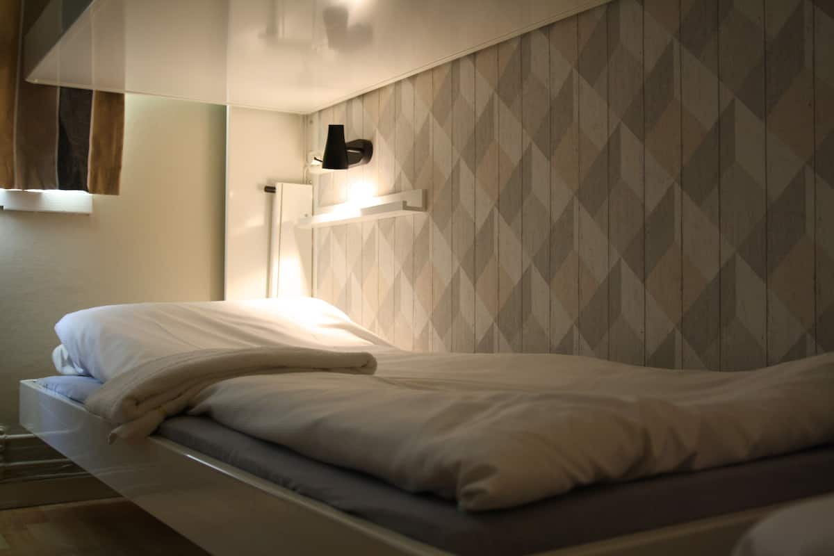 Cama bem arrumada com edredom branco e travesseiros, abajur e parede com padrão geométrico.