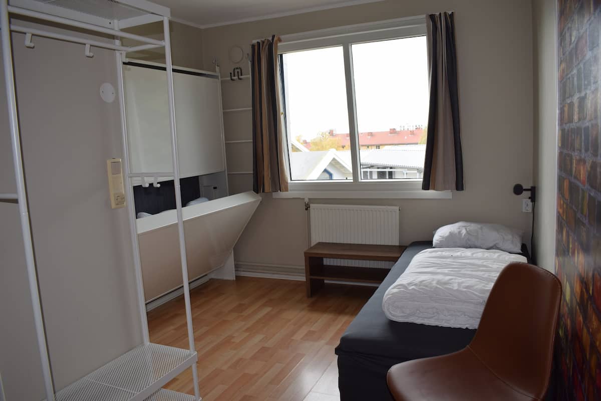 Quarto com cama de solteiro, uma cama individual, uma cadeira, janela com cortinas e radiador.