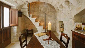 Interior rústico de pedra com mesa de jantar de madeira, vela e escada para um andar superior.