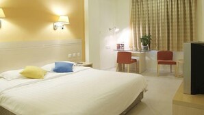 Quarto de hotel com cama, escrivaninha, duas cadeiras, televisão e janela com cortinas.