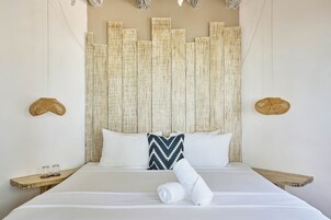 Quarto de hotel com uma cama grande, cabeceira texturizada, duas luminárias fixadas na parede e uma pequena mesa de madeira com copos.