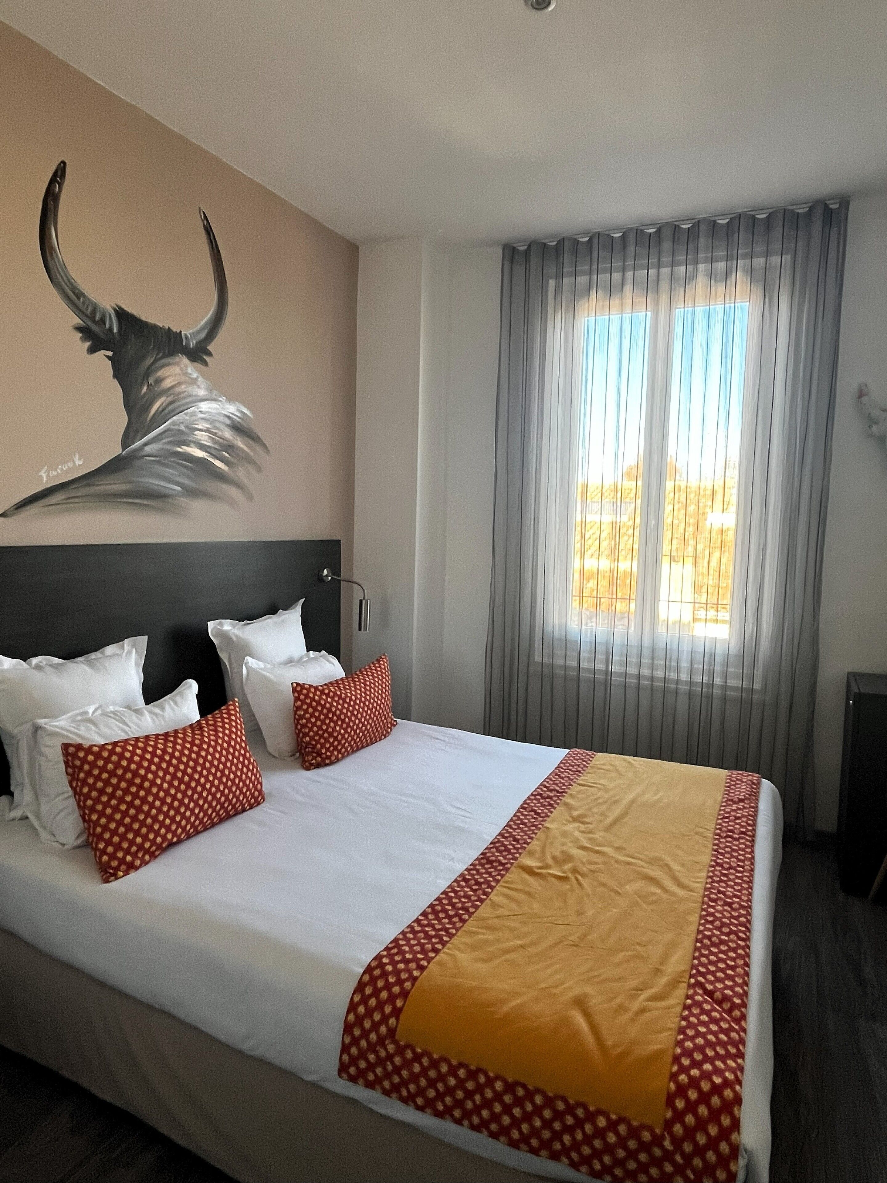 Quarto de hotel com uma cama grande, roupa de cama branca, almofadas de destaque vermelhas e amarelas, uma obra de arte na parede de cabeça de touro e uma janela com cortinas leves.