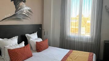 Quarto de hotel com uma cama grande, roupa de cama branca, almofadas de destaque vermelhas e amarelas, uma obra de arte na parede de cabeça de touro e uma janela com cortinas leves.