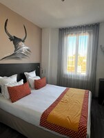 Quarto de hotel com uma cama grande, roupa de cama branca, almofadas de destaque vermelhas e amarelas, uma obra de arte na parede de cabeça de touro e uma janela com cortinas leves.