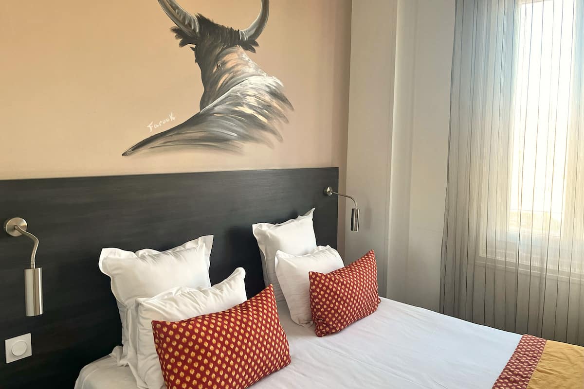 Quarto de hotel com cama, almofadas brancas, uma almofada vermelha estampada, uma pintura de chifre na parede e uma janela com cortinas.