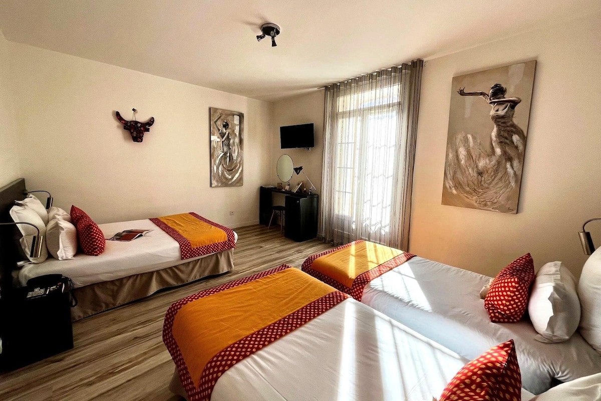 Quarto de hotel com duas camas, uma escrivaninha, uma cadeira, televisão e obras de arte nas paredes.