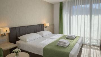 Quarto de hotel com uma cama, duas luminárias de cabeceira, uma mesa redonda com um vaso de flores e vista para o exterior através de cortinas finas.
