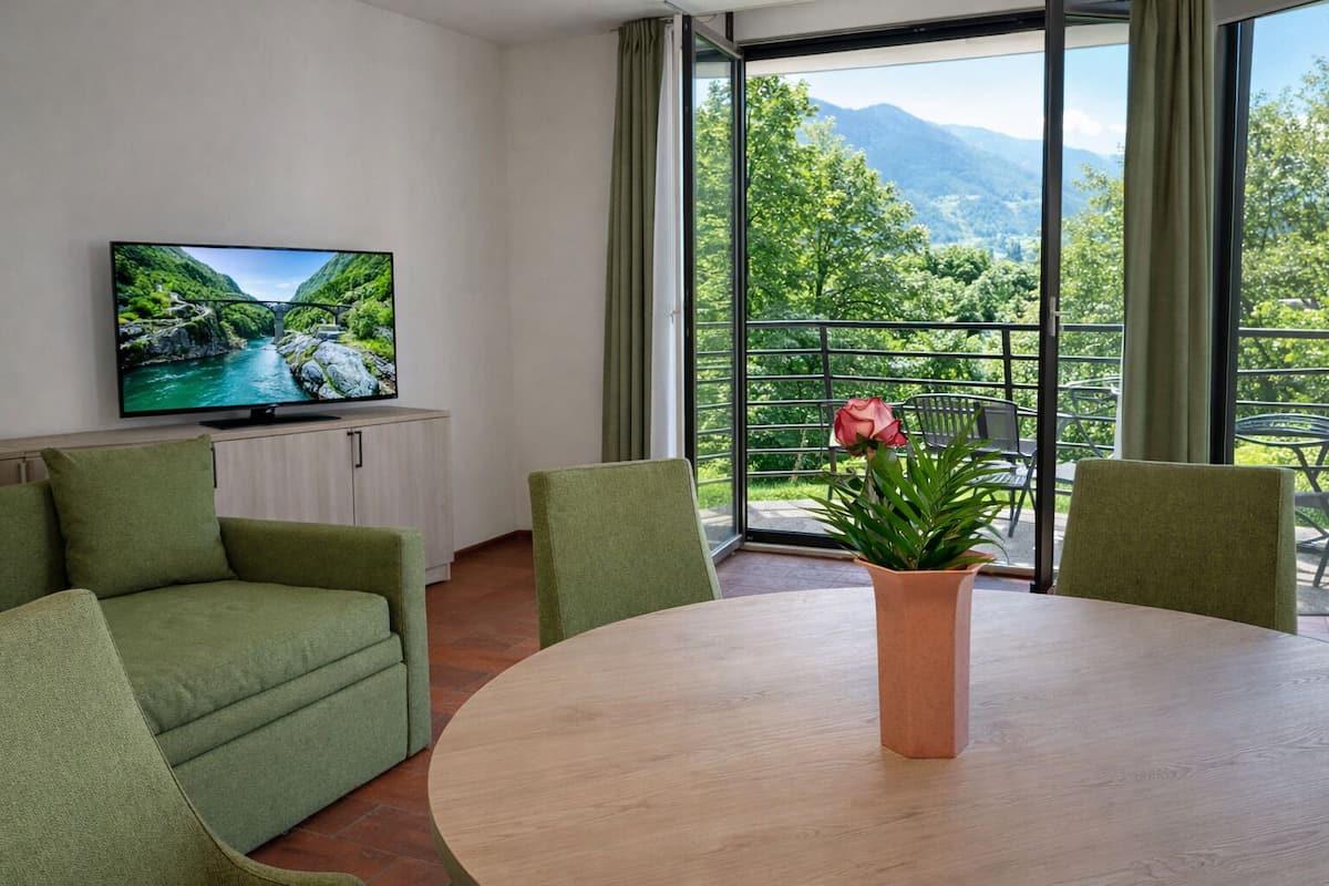 Quarto moderno com televisão em um armário, mesa redonda com cadeiras, planta em vaso e varanda com vista para vegetação e montanhas.