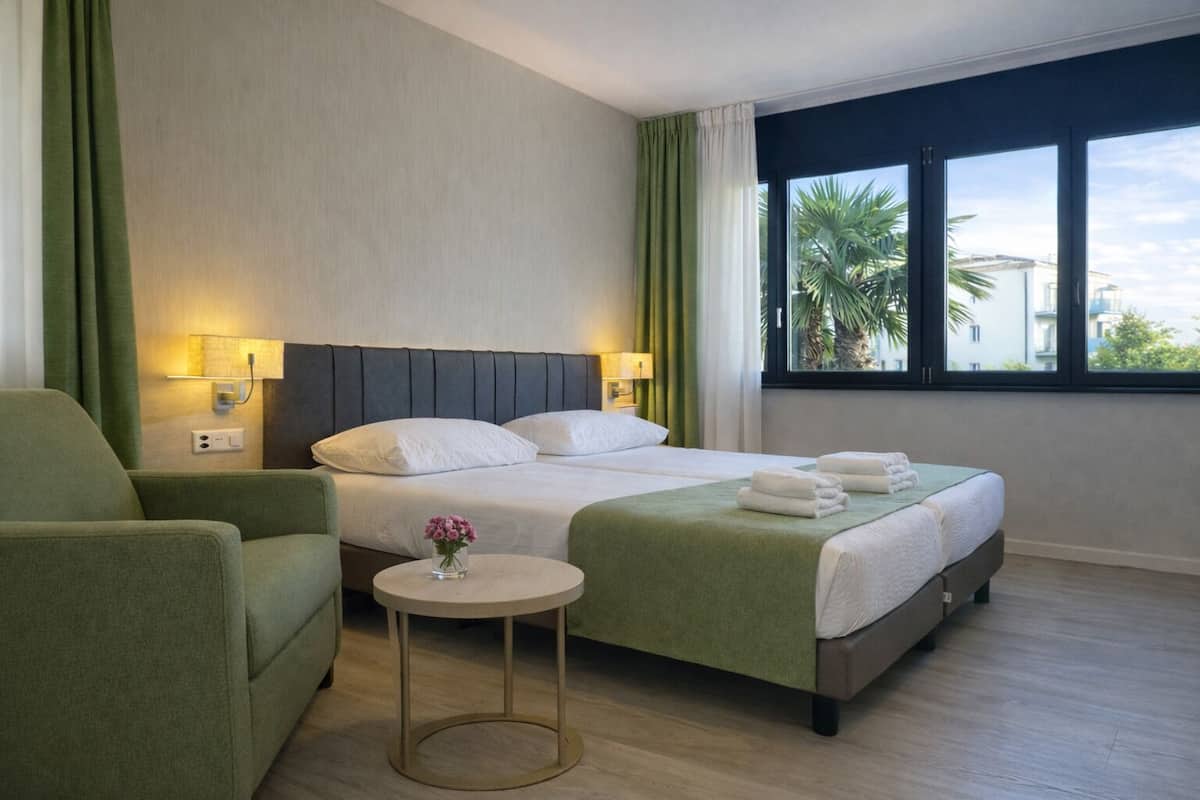Quarto de hotel com cama, duas mesas de cabeceira, uma cadeira e vista para palmeiras pela janela.