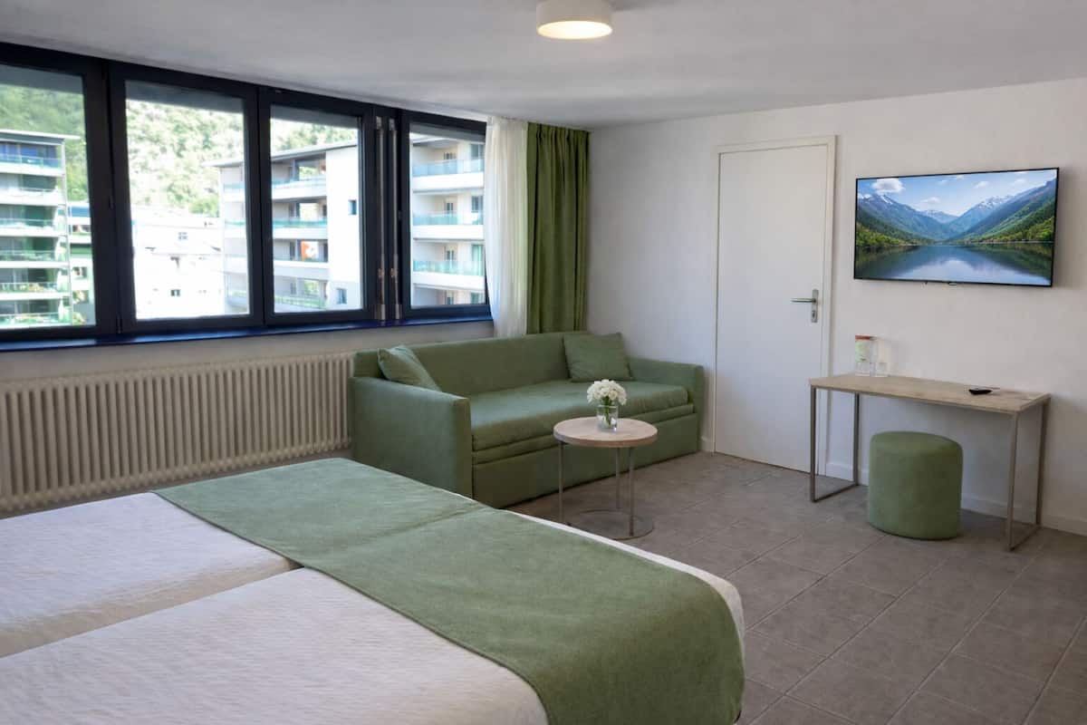 Quarto de hotel com uma cama, um sofá verde, uma pequena mesa redonda com um vaso de flores, uma escrivaninha com um computador e uma grande janela com vista para edifícios e vegetação.