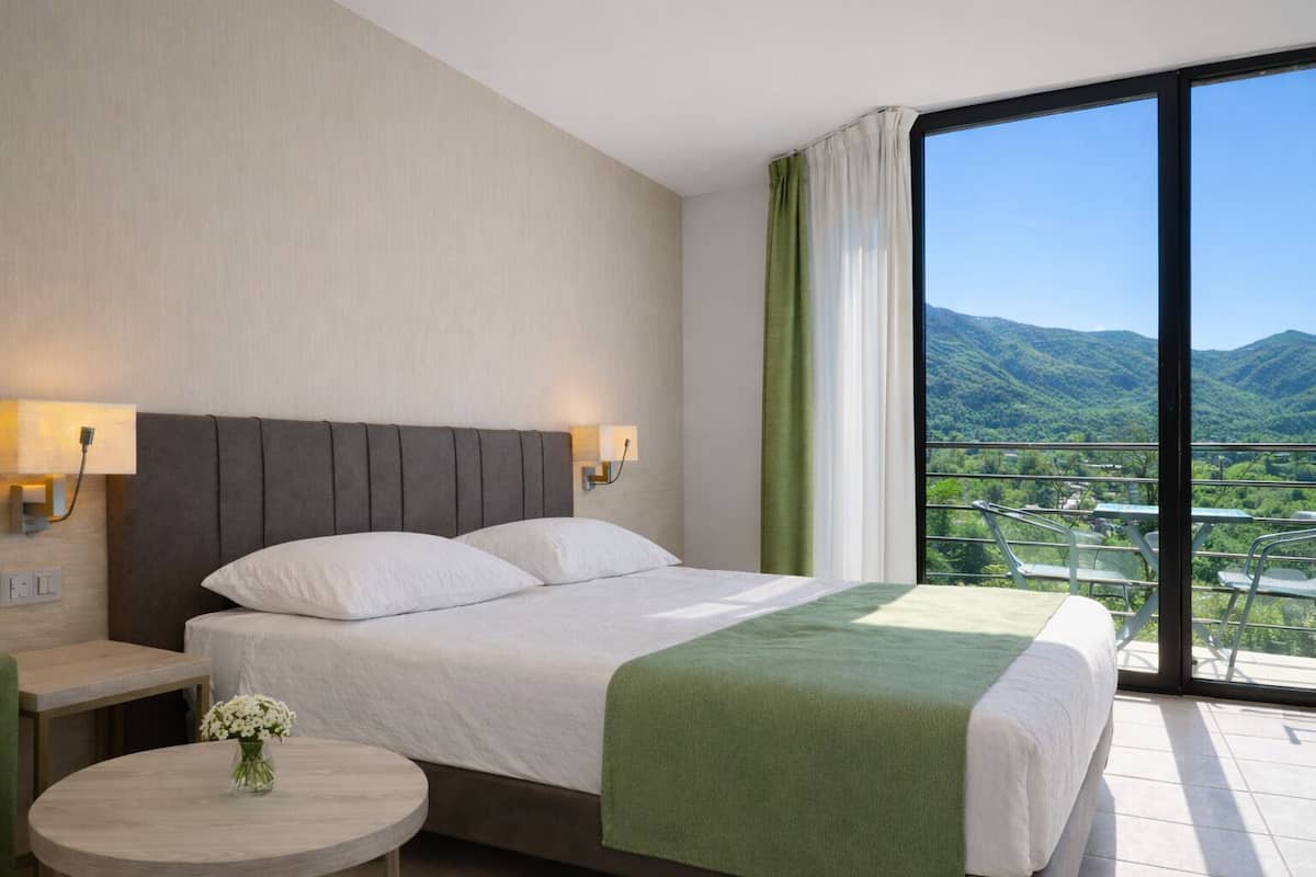 Quarto de hotel com uma cama, mesas de cabeceira, uma mesinha redonda com um vaso de flores e uma grande janela com vista para as montanhas.