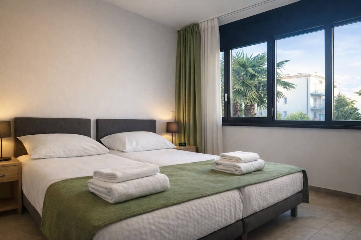 Uma cama de casal com colcha verde e duas almofadas brancas, uma mesinha de cabeceira com abajur e uma janela com vista para palmeiras e edifícios.