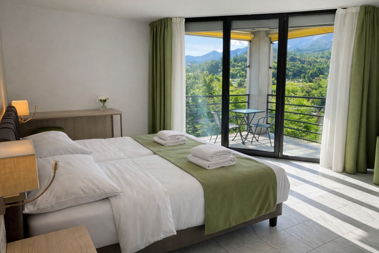 Quarto de hotel com uma cama, mesa de cabeceira, luminária e vista para uma varanda com mesa e cadeiras.
