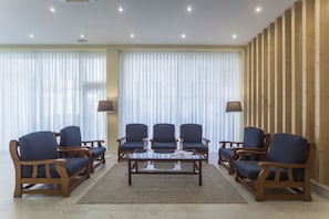 Uma sala de conferências com seis cadeiras de madeira e uma mesa de vidro, janelas amplas com persianas e lambrisas de madeira em uma parede.