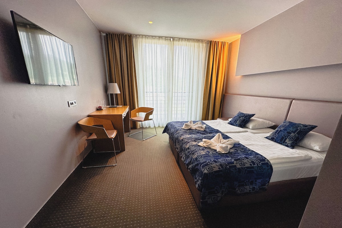 Quarto de hotel com cama, duas cadeiras, escrivaninha e uma janela grande com cortinas.