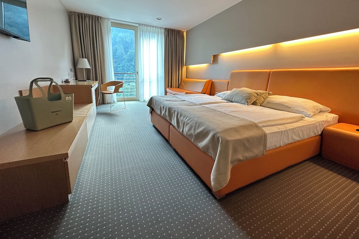 Quarto de hotel com cama, mesas de cabeceira, escrivaninha, cadeira e vista para o exterior.
