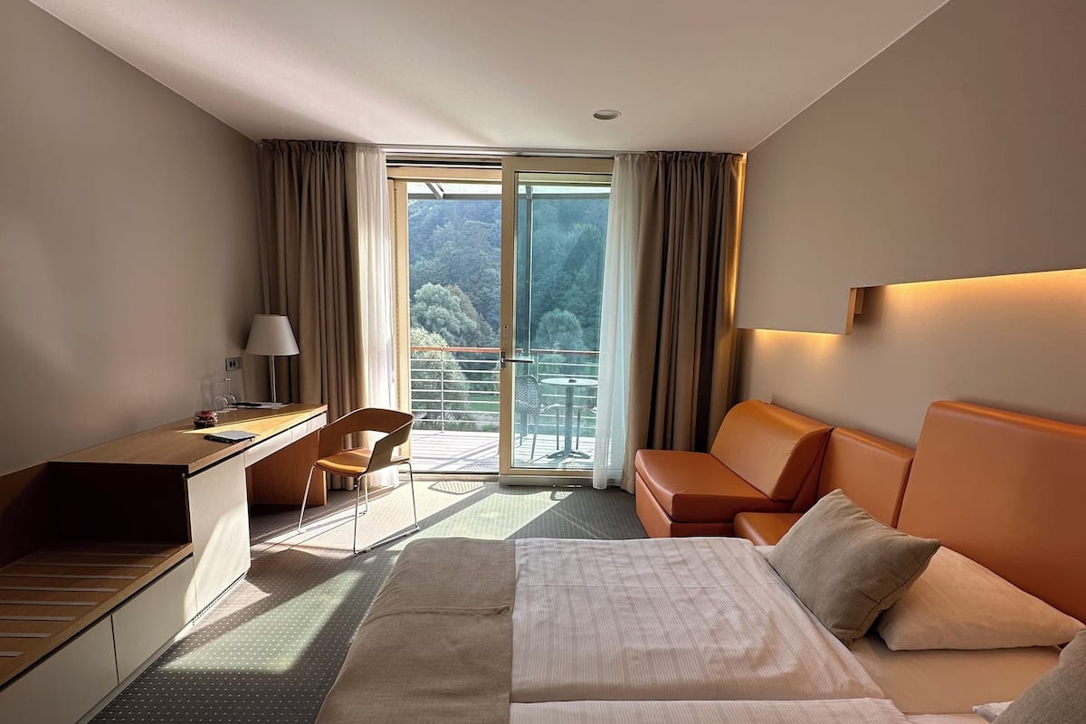 Quarto de hotel com cama, escrivaninha, cadeira e varanda com vista para a floresta.