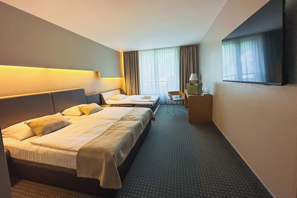 Quarto de hotel com duas camas, uma escrivaninha, uma cadeira, um abajur e uma janela grande com cortinas.
