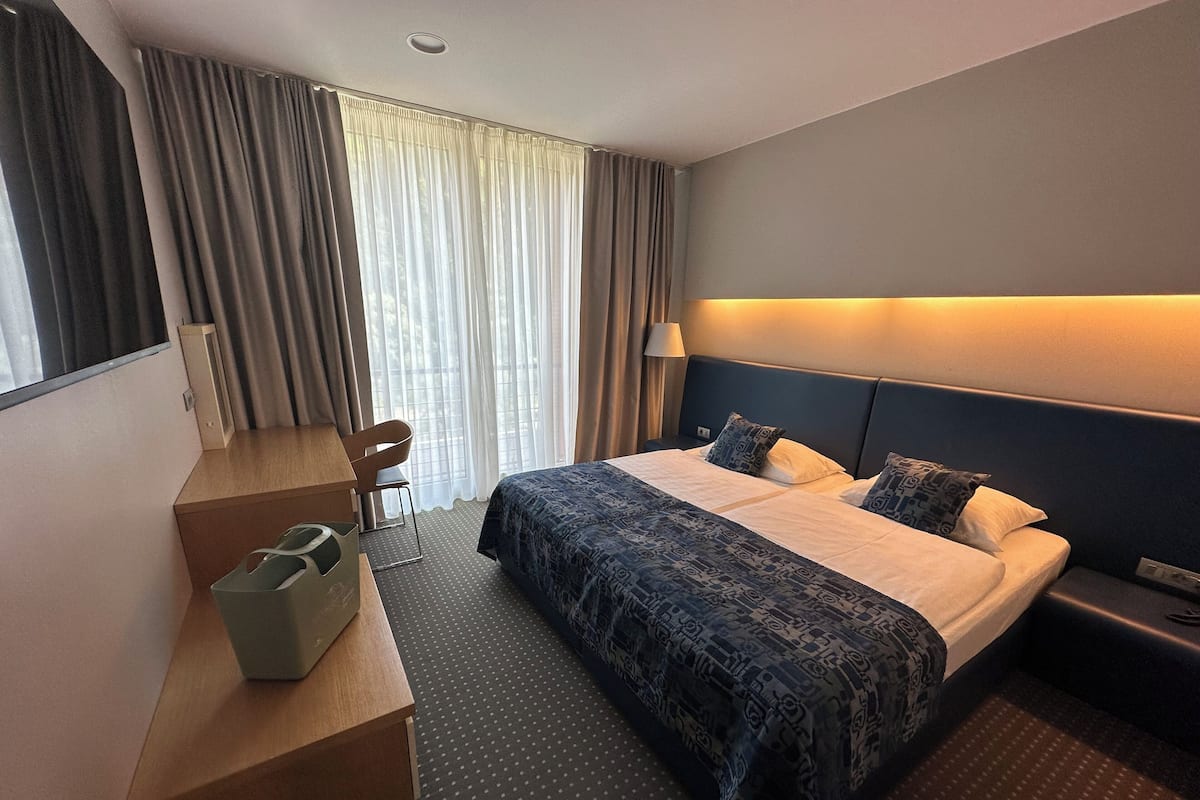 Quarto de hotel com uma cama, uma escrivaninha, uma cadeira e uma janela com cortinas.