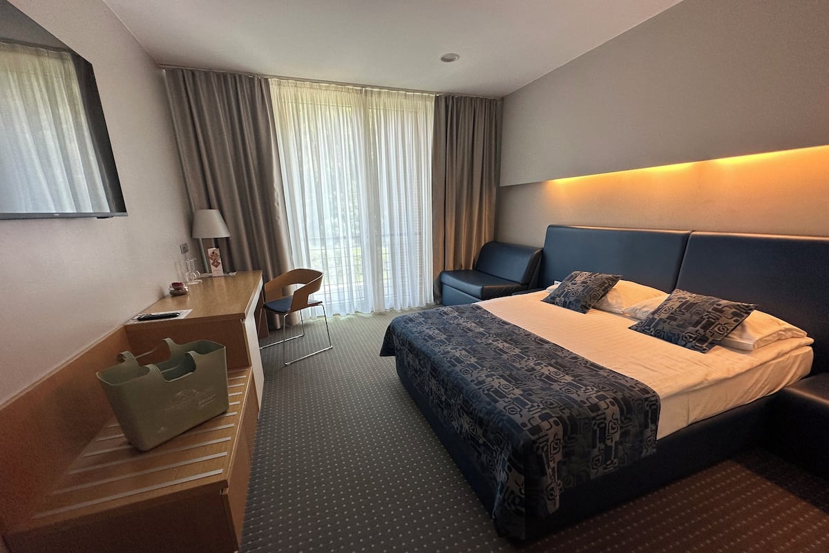 Quarto de hotel com cama, escrivaninha, cadeira e janela com cortinas.