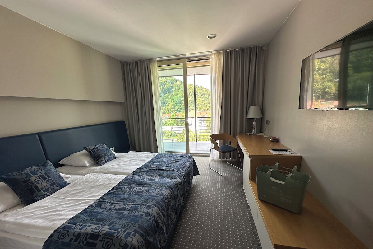 Quarto de hotel com cama, escrivaninha, cadeira e vista para áreas verdes.