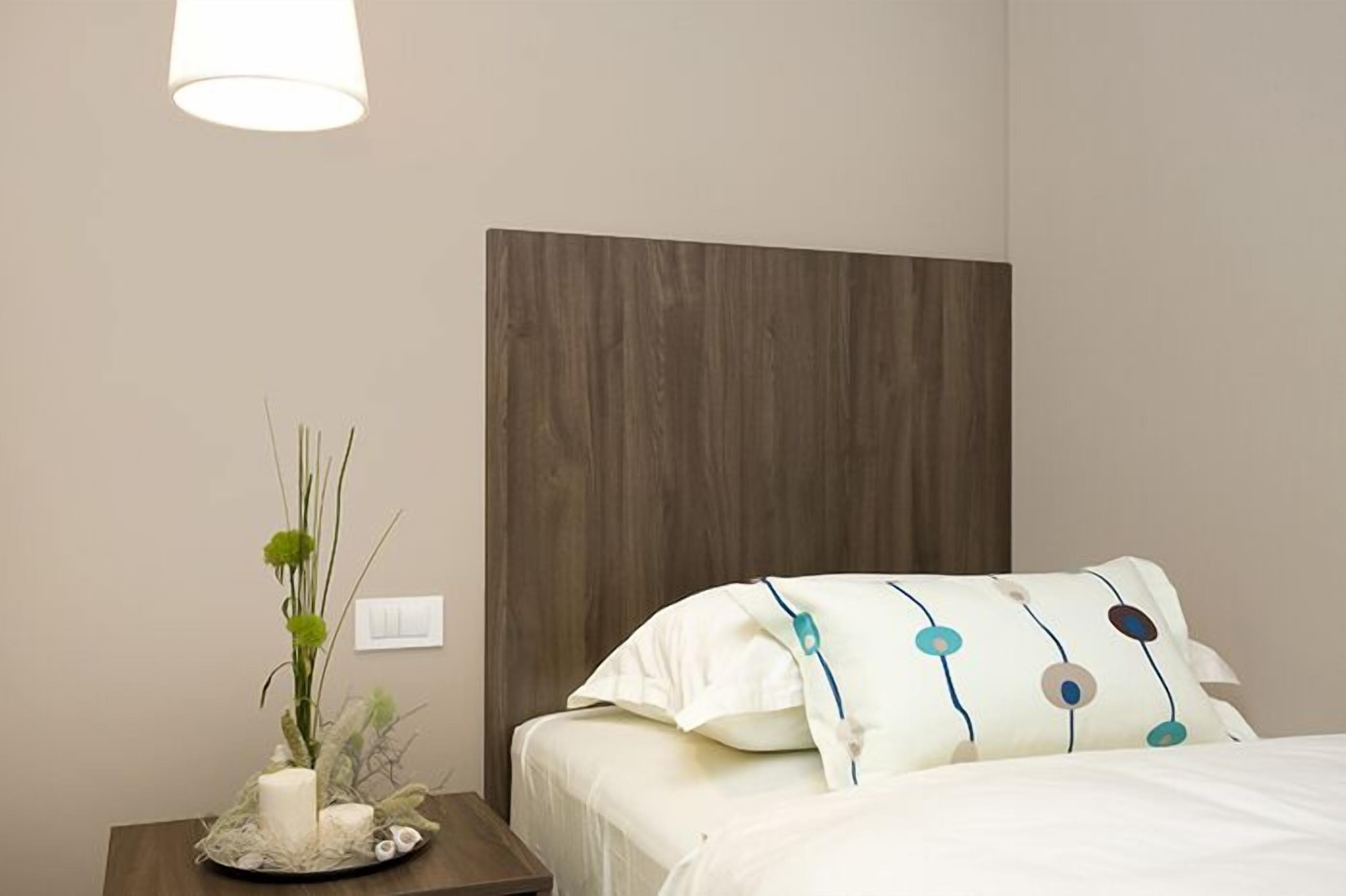 Quarto com cabeceira de madeira, cama branca com almofadas estampadas, mesa de cabeceira com um vaso de plantas e abajur branco.