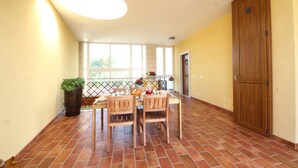 Um quarto amplo com piso de azulejos, uma mesa de jantar com cadeiras, uma grande porta de madeira e vista pela janela.