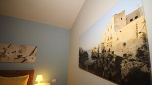 Quarto com cama, mesa de cabeceira, abajur e um grande quadro na parede retratando um edifício histórico.