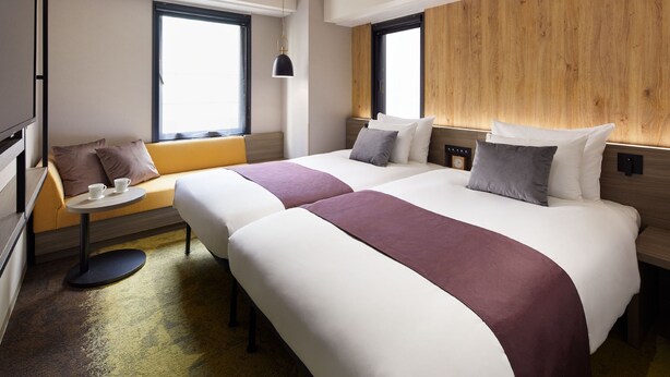 Quarto de hotel com duas camas, um sofá, uma pequena mesa com xícaras e janelas amplas.