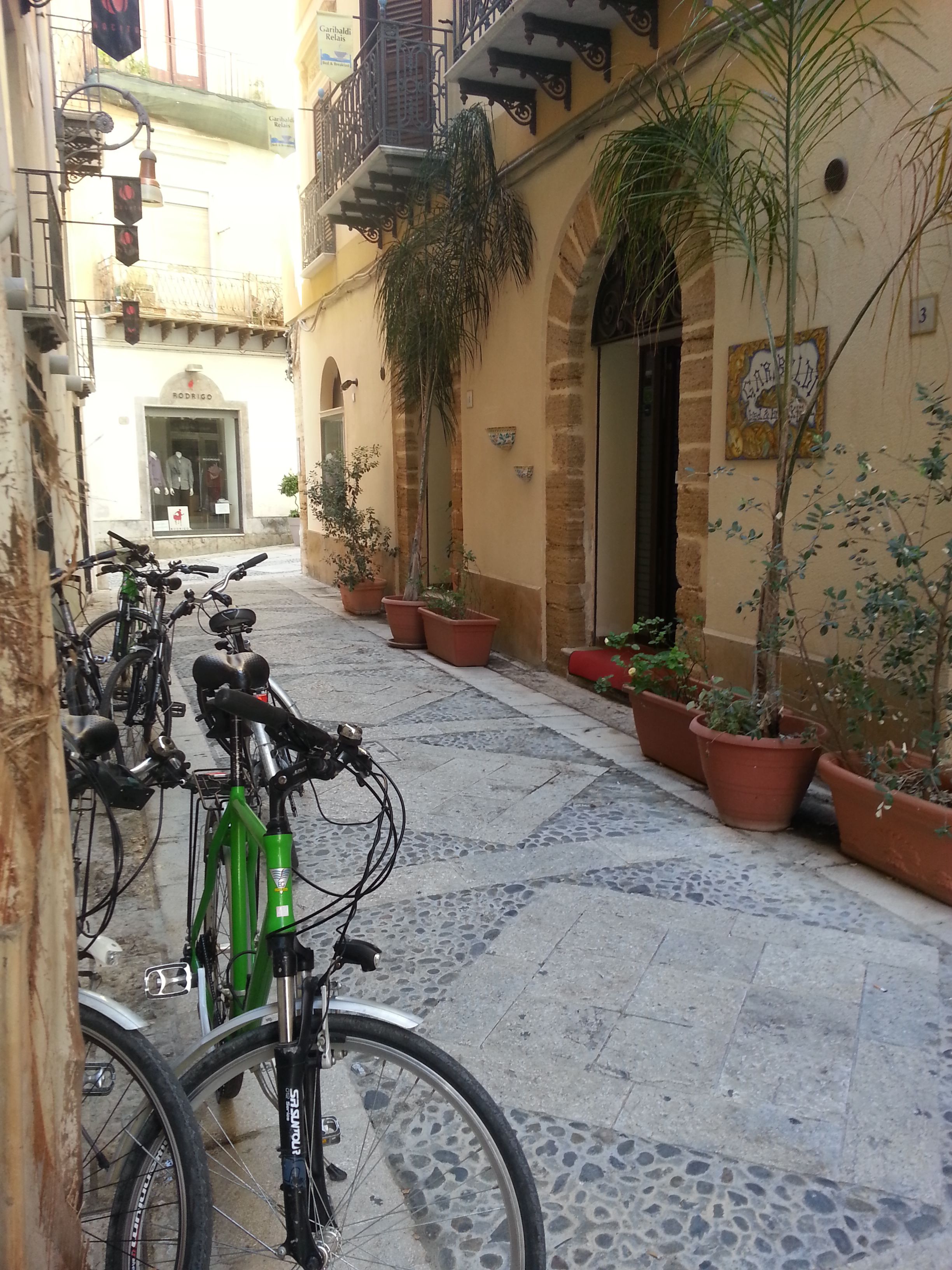 Uma rua estreita com bicicletas estacionadas na lateral, plantas em vasos ao longo do caminho e um prédio com uma placa indicando 